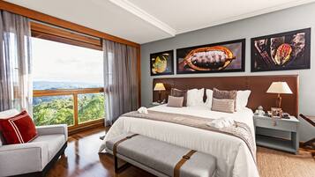 Quarto de hotel com uma cama grande, um banquinho, um sofá e uma mesinha de cabeceira. Há três quadros na parede.