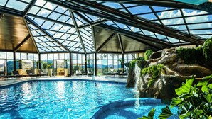 Piscina interior com teto de vidro, espreguiçadeiras e vista para as montanhas.