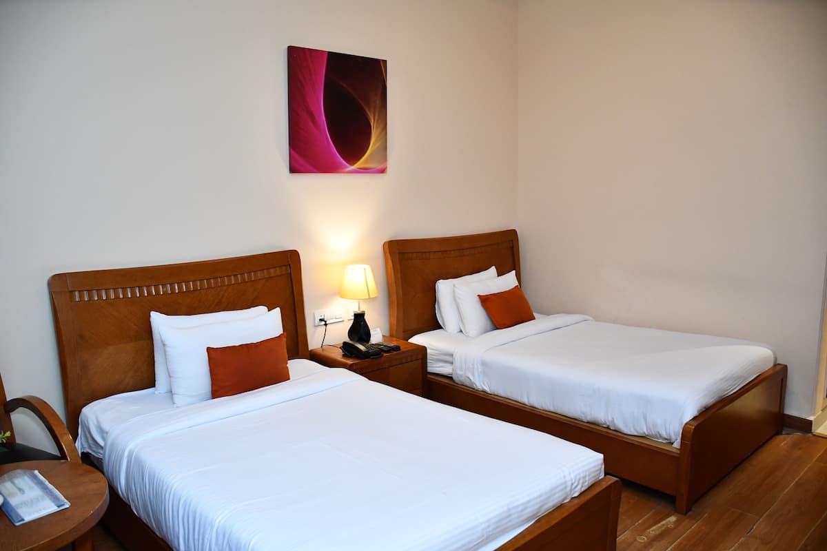 Duas camas de solteiro com cabeceiras de madeira, roupa de cama branca e almofadas laranja. Há uma mesa de cabeceira com uma luminária e um telefone. Um quadro está visível na parede acima das camas.