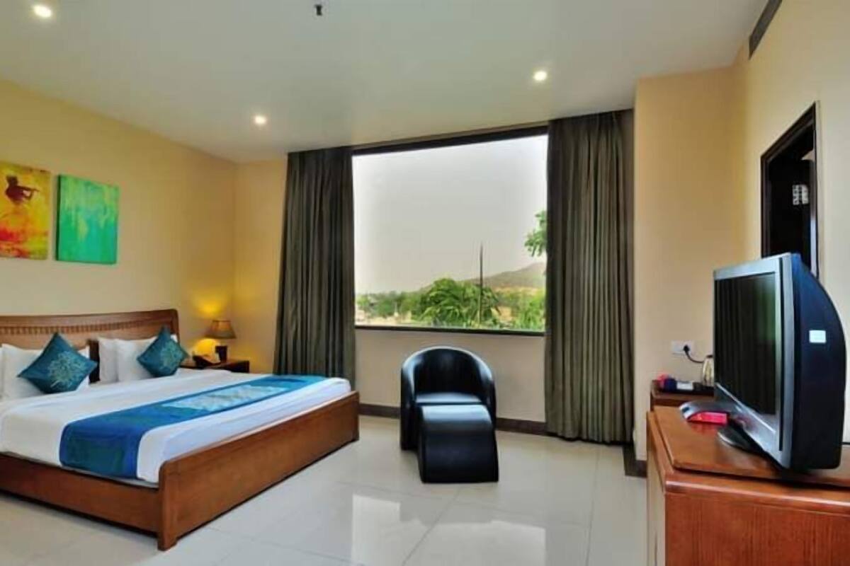 Quarto de hotel com cama, televisão, escrivaninha e janela com vista para o verde.