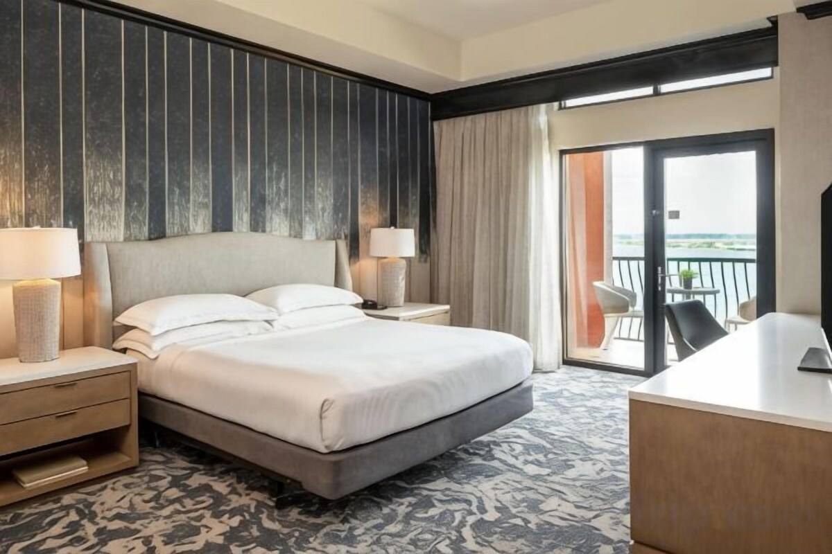 Quarto de hotel com cama, mesas de cabeceira, escrivaninha e varanda com vista.