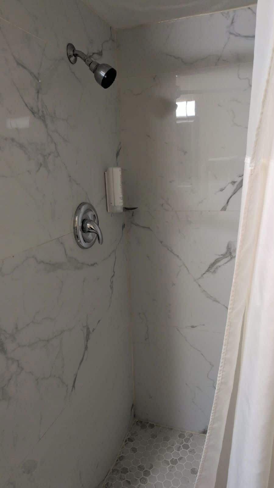 Chuveiro com chuveiro manual, comando fixado na parede e chão revestido com azulejos.