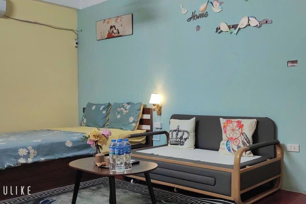 Quarto com cama, sofá, mesa pequena com abajur e janela com cortinas.