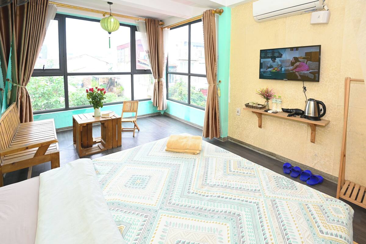 Quarto de hotel com uma cama, uma mesa, uma cadeira, uma janela com cortinas, uma televisão e uma estante com objetos.