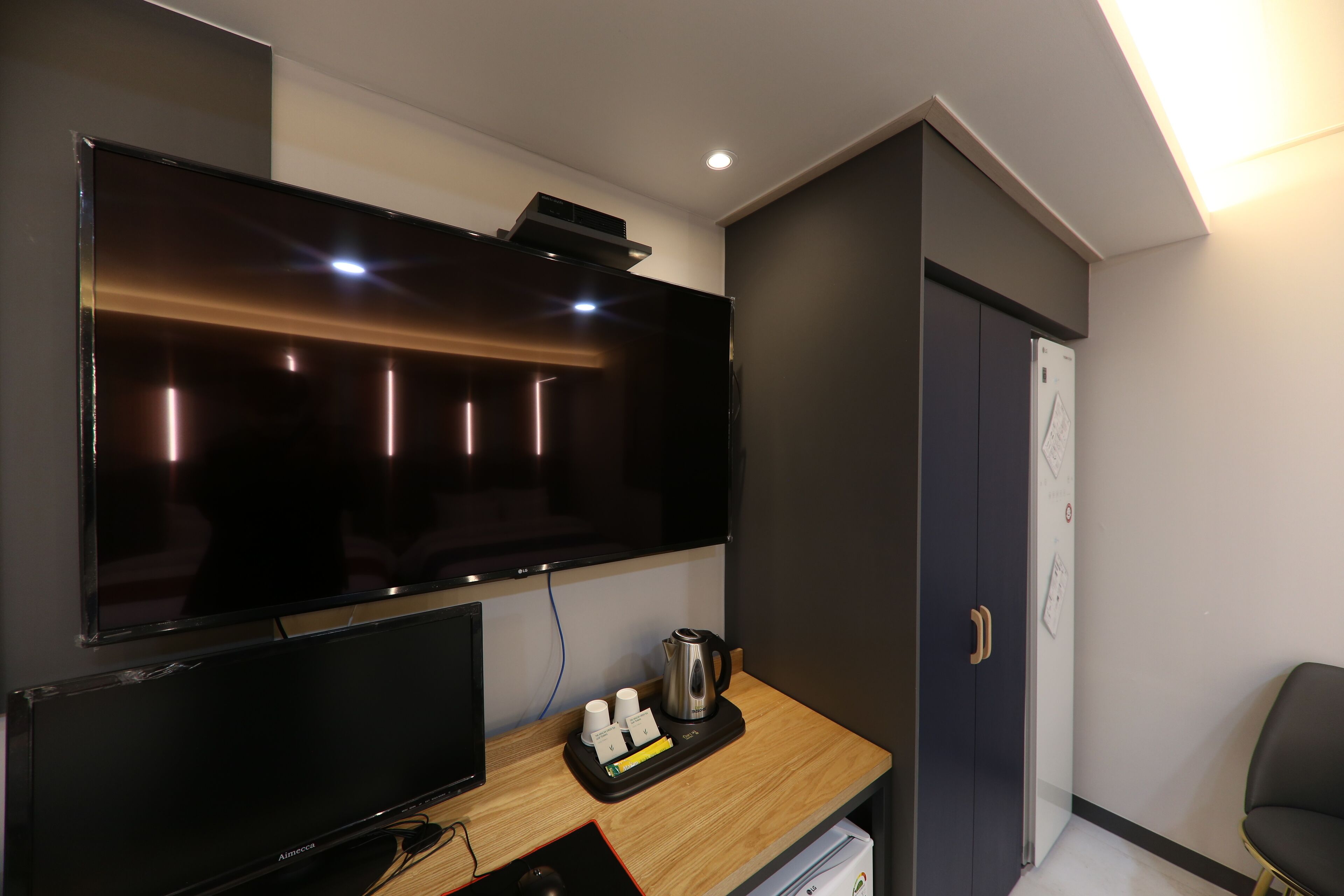 Quarto de hotel moderno com uma grande TV de tela plana na parede, uma escrivaninha de madeira com chaleira e xícaras, e um guarda-roupa de cor escura.