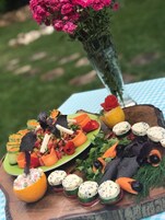 Uma tábua de madeira com diversos alimentos, incluindo uma salada, cubos de queijo e um arranjo floral decorativo.
