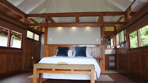 Quarto com estrutura de madeira, uma cama grande, uma mesa pequena e uma janela.