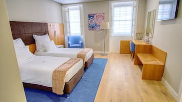 Quarto de hotel com duas camas, uma escrivaninha, uma cadeira, um abajur e uma janela grande.