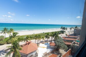 Vista da praia com céu claro, palmeiras e um edifício com telhado vermelho.