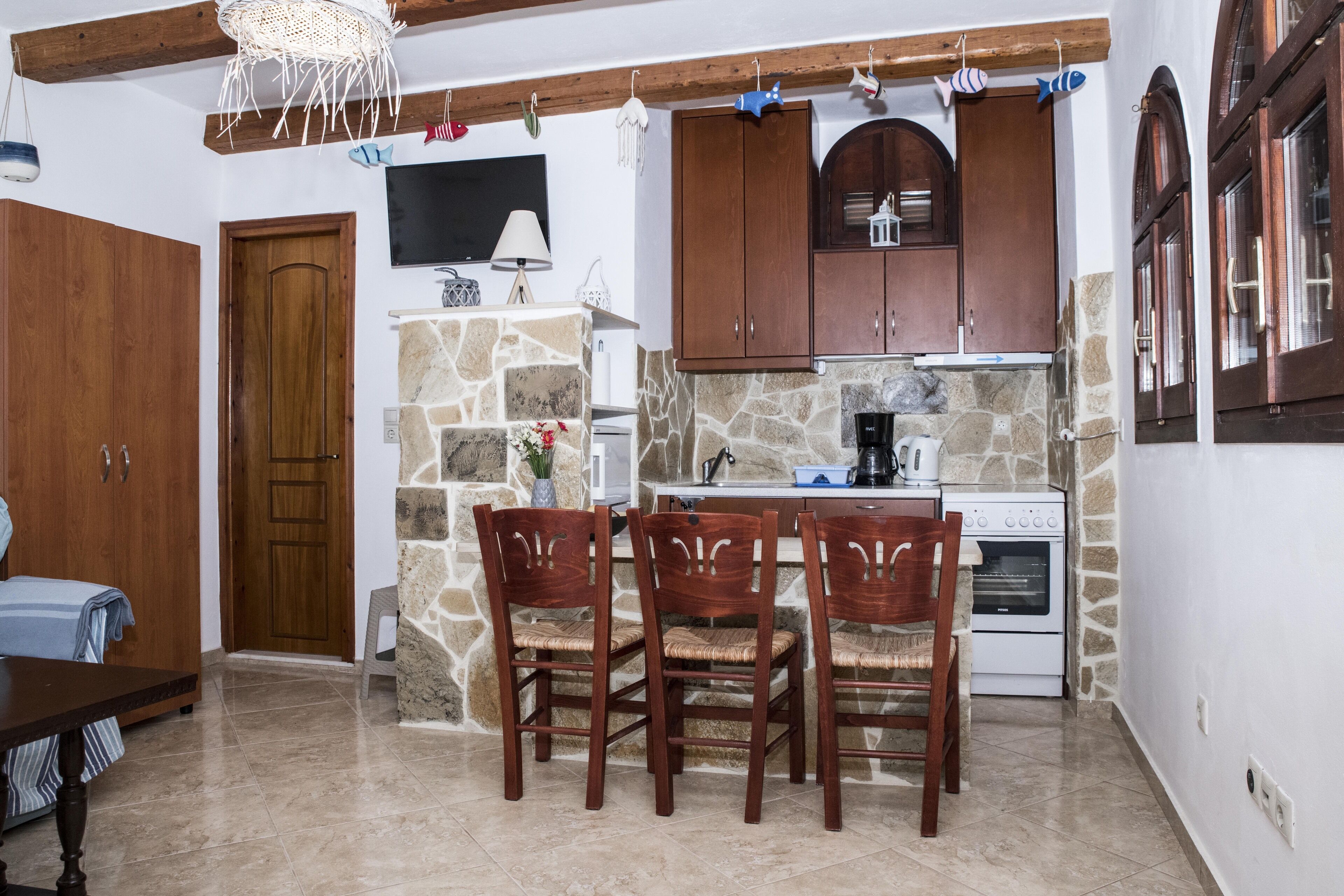 Uma cozinha com armários de madeira, parede de pedra, mesa de jantar com cadeiras e uma TV fixada acima dos armários.