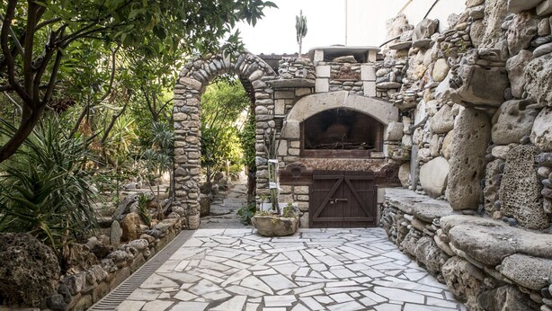Um caminho de pedra leva a um forno de pedra com design rústico, cercado por vegetação e muros de pedra.
