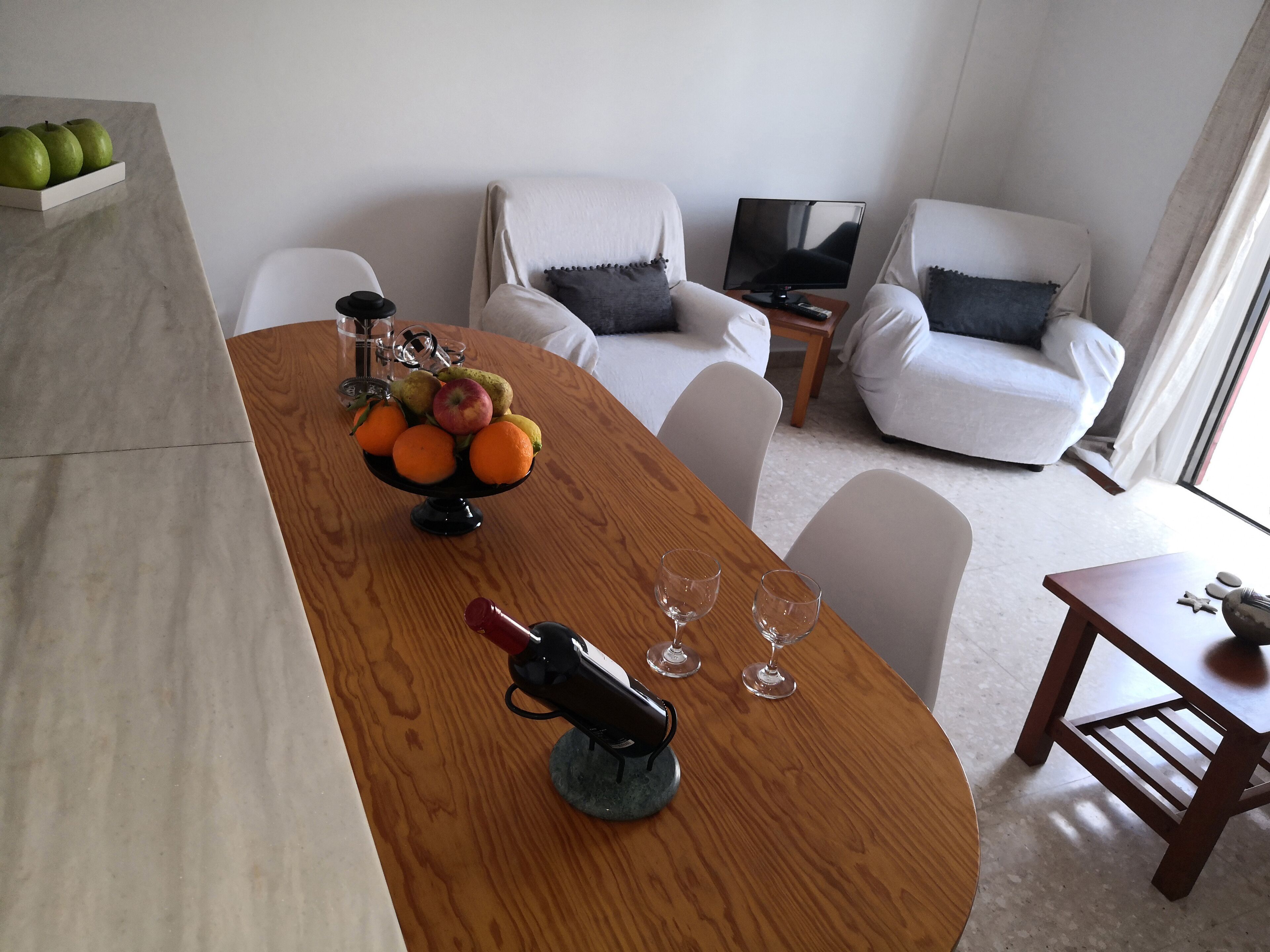 Uma sala de estar moderna com uma mesa de jantar de madeira, uma cesta de frutas, duas taças de vinho e uma garrafa de vinho. Há dois sofás brancos com almofadas cinzentas, uma televisão e uma pequena mesa lateral de madeira.