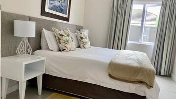 Um quarto com cama, mesa de cabeceira, luminária, quadro pendurado na parede e janela com cortinas.