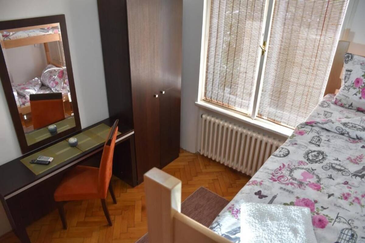 Quarto com escrivaninha de madeira, cadeira, cama com roupa de cama florida, janela com persianas e radiador.