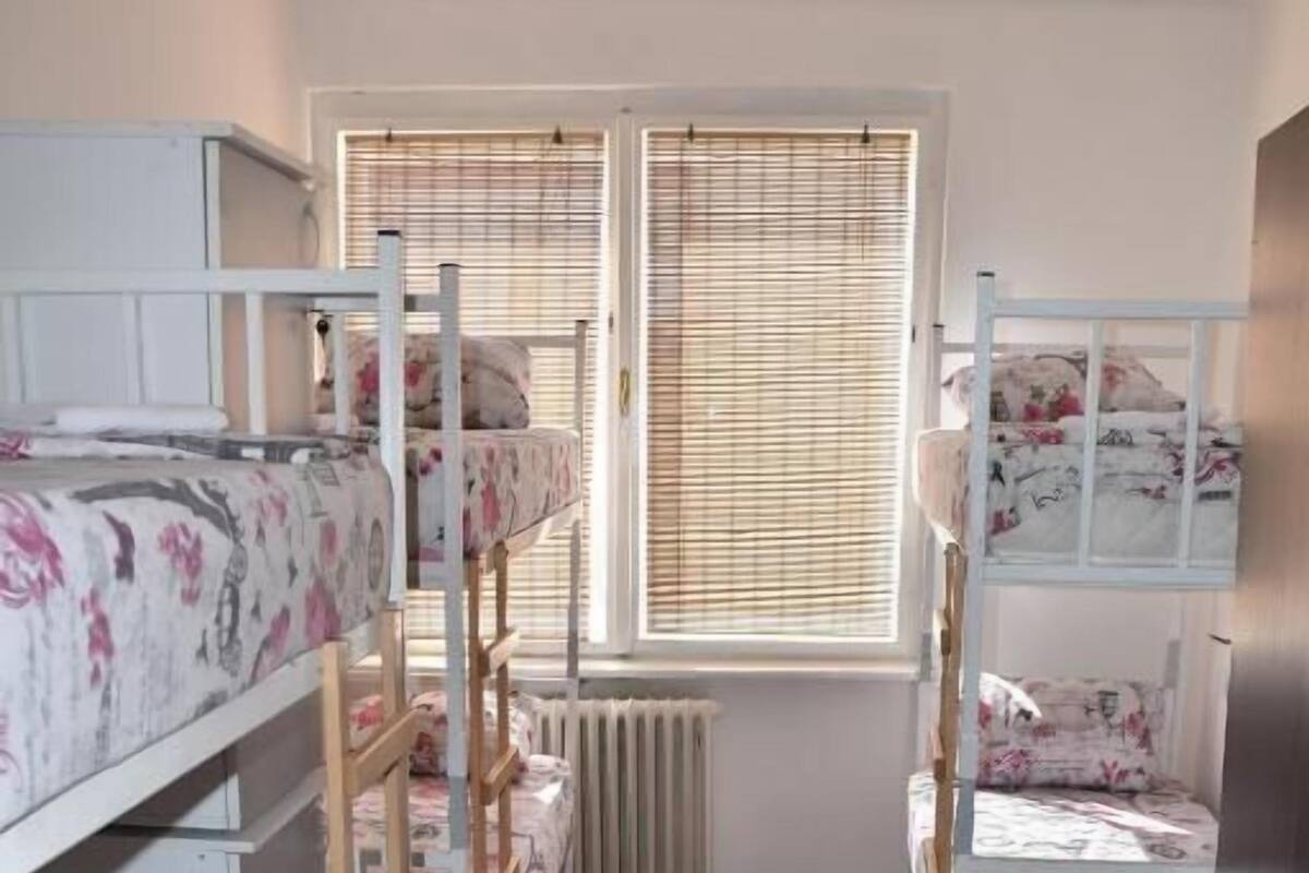 Um quarto de estudante com beliches, um radiador, uma janela com persianas e piso coberto de carpete.
