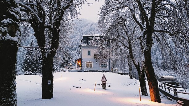 Paisagem coberta de neve com uma casa de vários andares, uma árvore carregada de neve e uma cerca também coberta de neve.