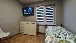 Quarto com cama, televisão na parede, janela com persianas e cômoda branca.