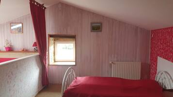 Quarto com teto inclinado, janela, radiador, cama com cobertura vermelha, cadeira e pequena mesa com um vaso de flores rosa.