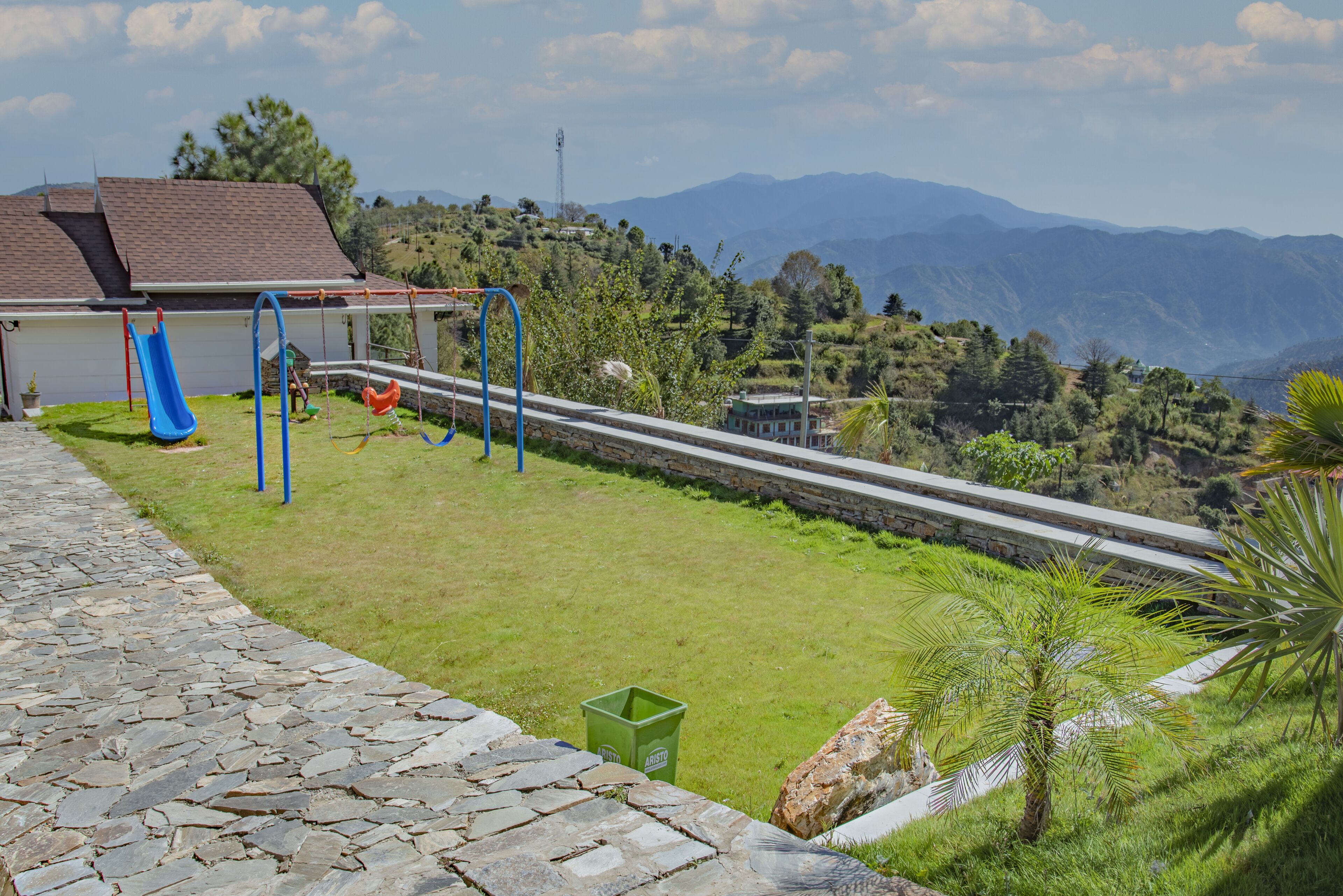 Uma trilha de pedra leva a uma casa com escorrega e balanço azuis, cercada por vegetação exuberante e montanhas ao fundo.