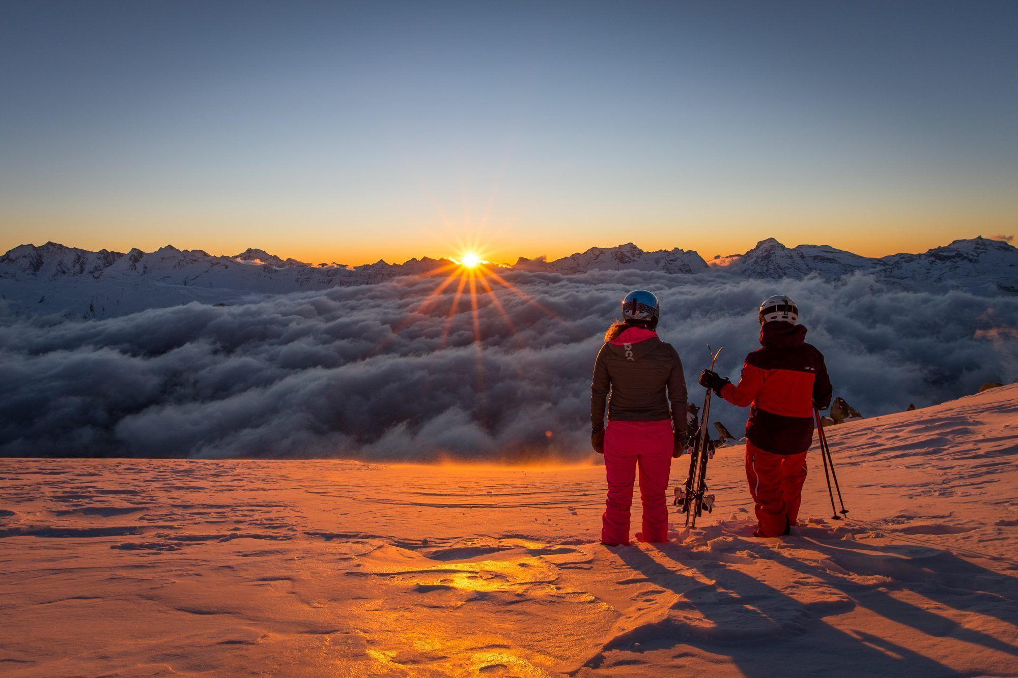 Duas pessoas em uma montanha nevada apreciando o pôr do sol atrás das nuvens.