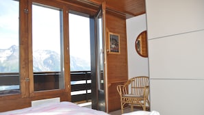 Um quarto com cama, uma cadeira de vime, janela com vista para as montanhas e um quadro na parede.
