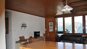 Ampla sala de estar com uma mesa de madeira, televisão e sofá. Janela com vista para as montanhas.