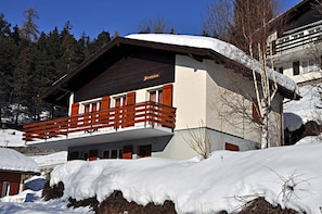 Um chalé de madeira com varanda, rodeado por neve na terra e árvores.