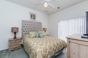 Quarto com cabeceira estofada, cama com roupa de cama estampada, duas luminárias de cabeceira, mesa de cabeceira, penteadeira, ventilador de teto e janela com persianas.