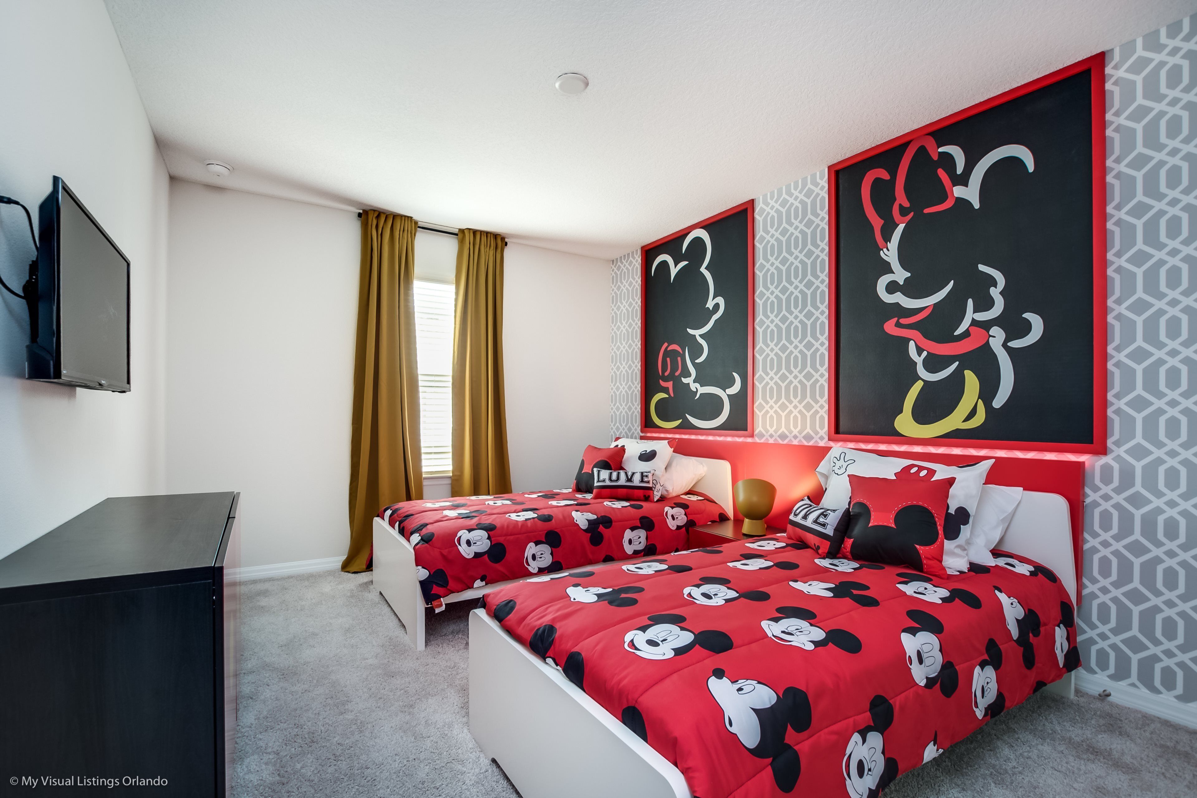Quarto de hotel com duas camas, cada uma coberta com roupa de cama vermelha com personagens de desenho animado, uma TV de tela plana e uma grande obra de arte na parede.