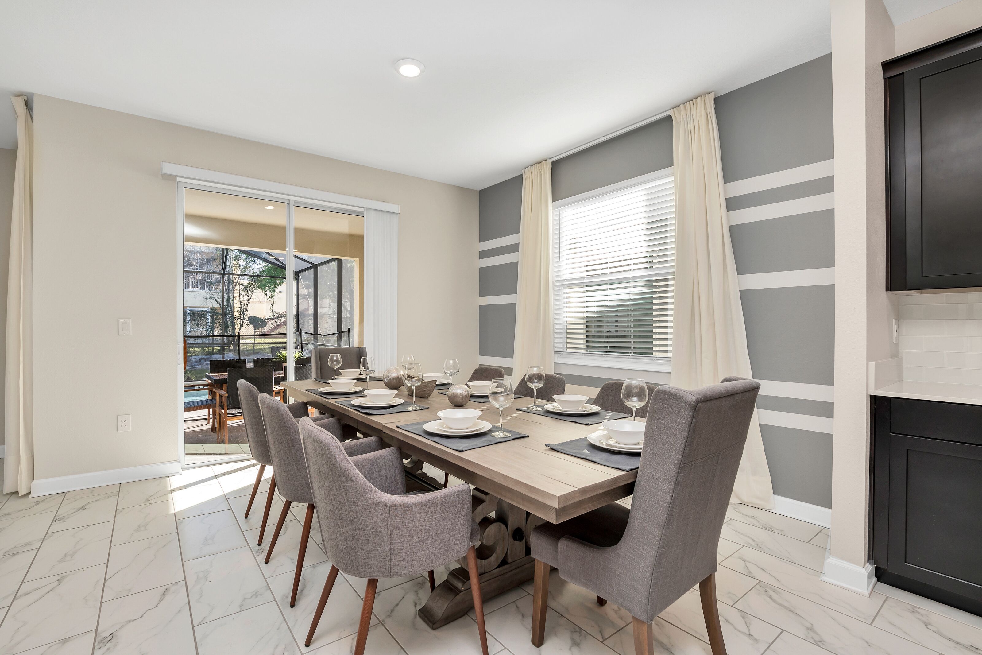 Uma sala de jantar com uma mesa comprida posta para seis pessoas, cadeiras cinzentas, uma lareira e vista pela janela com cortinas.