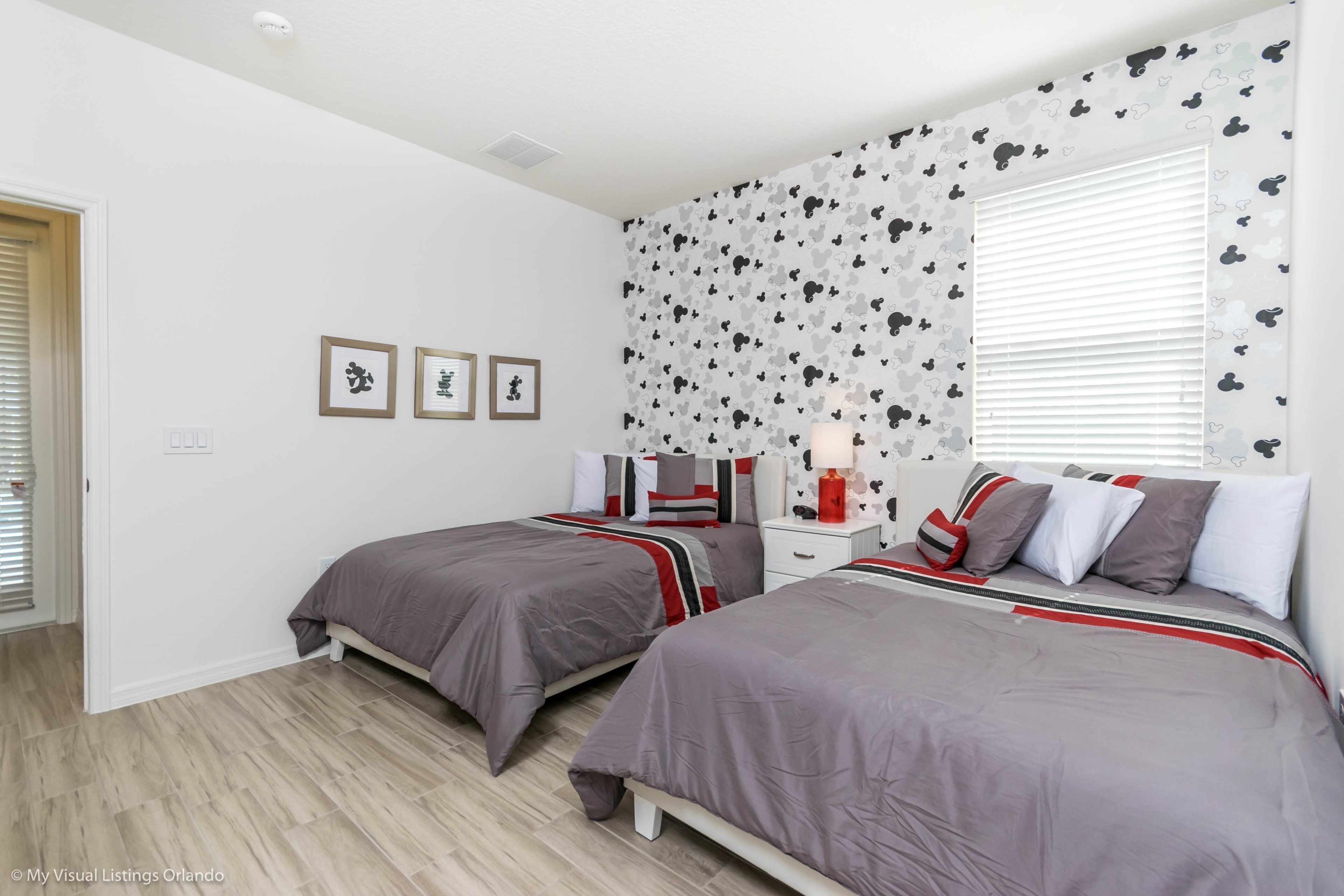 Quarto com duas camas, uma parede branca com padrão do Mickey Mouse, uma janela com persianas e uma mesa de cabeceira com abajur.