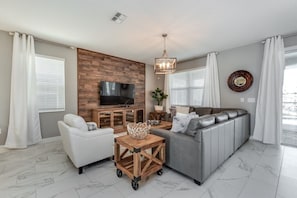 Uma sala de estar moderna com um sofá modular cinza, poltronas brancas, uma mesa de centro de madeira e uma TV fixada em uma parede de madeira.