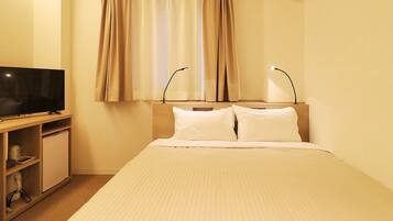 Quarto de hotel com uma cama, uma televisão sobre um suporte de madeira, uma mesa de trabalho com abajur e uma janela com cortinas.