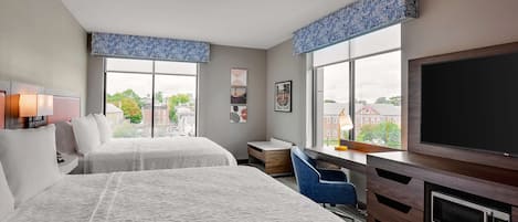 Quarto de hotel com duas camas, uma escrivaninha, uma televisão e grandes janelas com vista para a cidade.