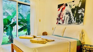 Quarto com cama, mesa de cabeceira, cadeira, janela com vista para o verde e um quadro na parede.