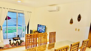 Uma sala de jantar com mesa de vidro e cadeiras de madeira, uma televisão sobre um suporte e vista para o exterior através de uma grande janela.