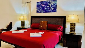 Quarto de hotel com uma cama grande, duas mesas de cabeceira com luminárias, um criado-mudo, uma cadeira e um quadro na parede.
