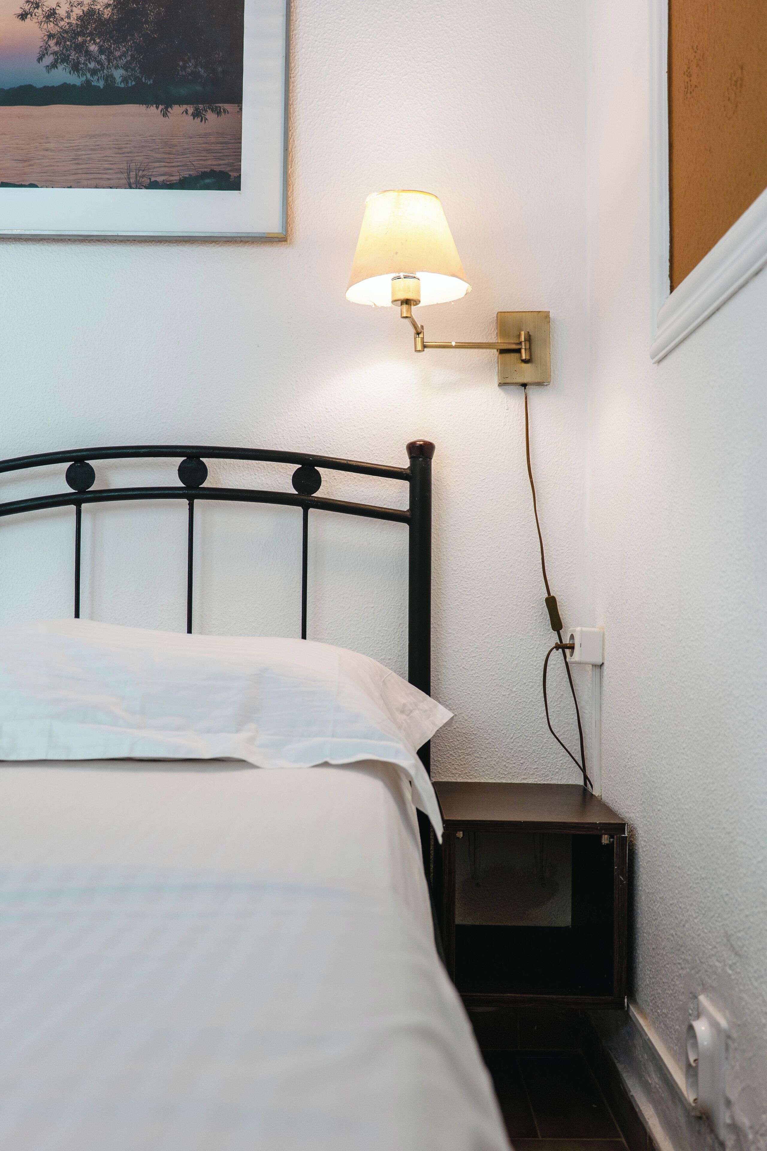 Uma cama bem arrumada com lençóis brancos, uma luminária na parede e uma mesinha de cabeceira.