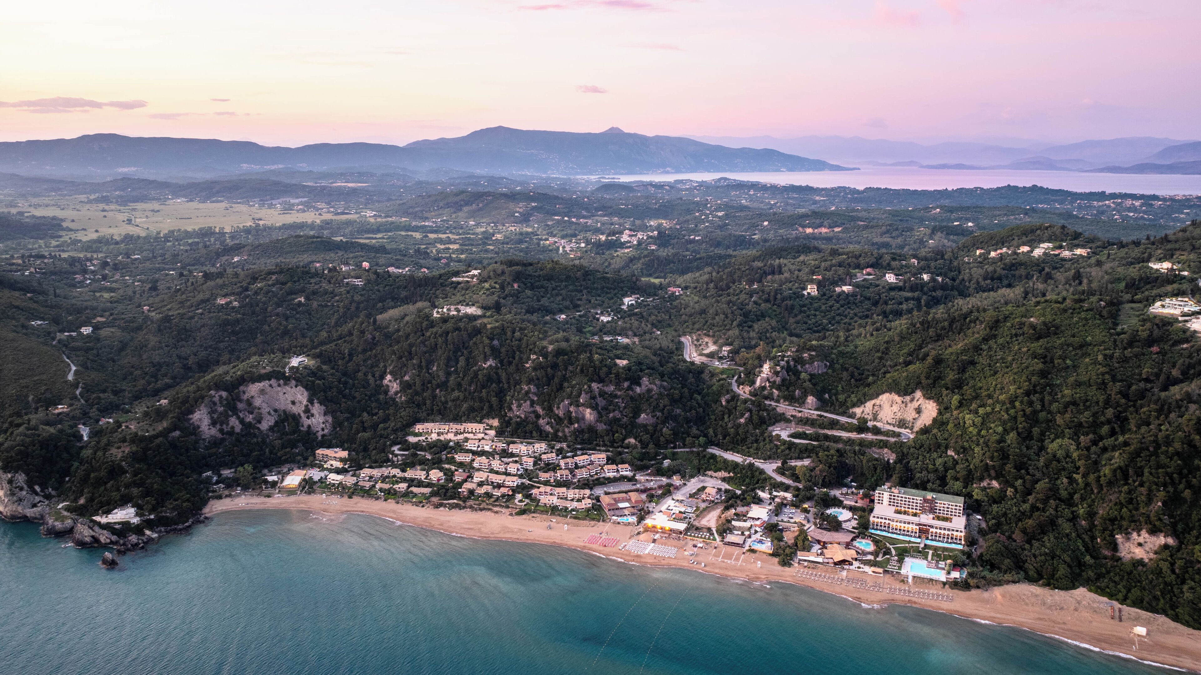 Vista aérea de uma área costeira com uma praia, um hotel grande com vários edifícios e colinas ao redor.