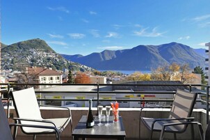 Uma varanda com mesa, cadeiras, garrafa e copos, com vista para uma cidade e montanhas.