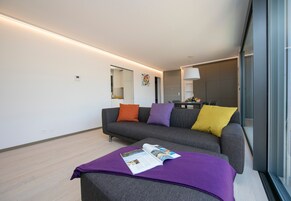 Uma sala de estar moderna com um sofá cinzento, almofadas coloridas, uma revista sobre um banquinho roxo e uma parede de vidro que dá para a cozinha.