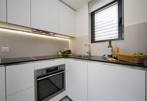 Cozinha moderna com armários brancos, forno embutido, exaustor de aço inoxidável, janela com persianas e bancada com uma garrafa de vinho, legumes e pão.