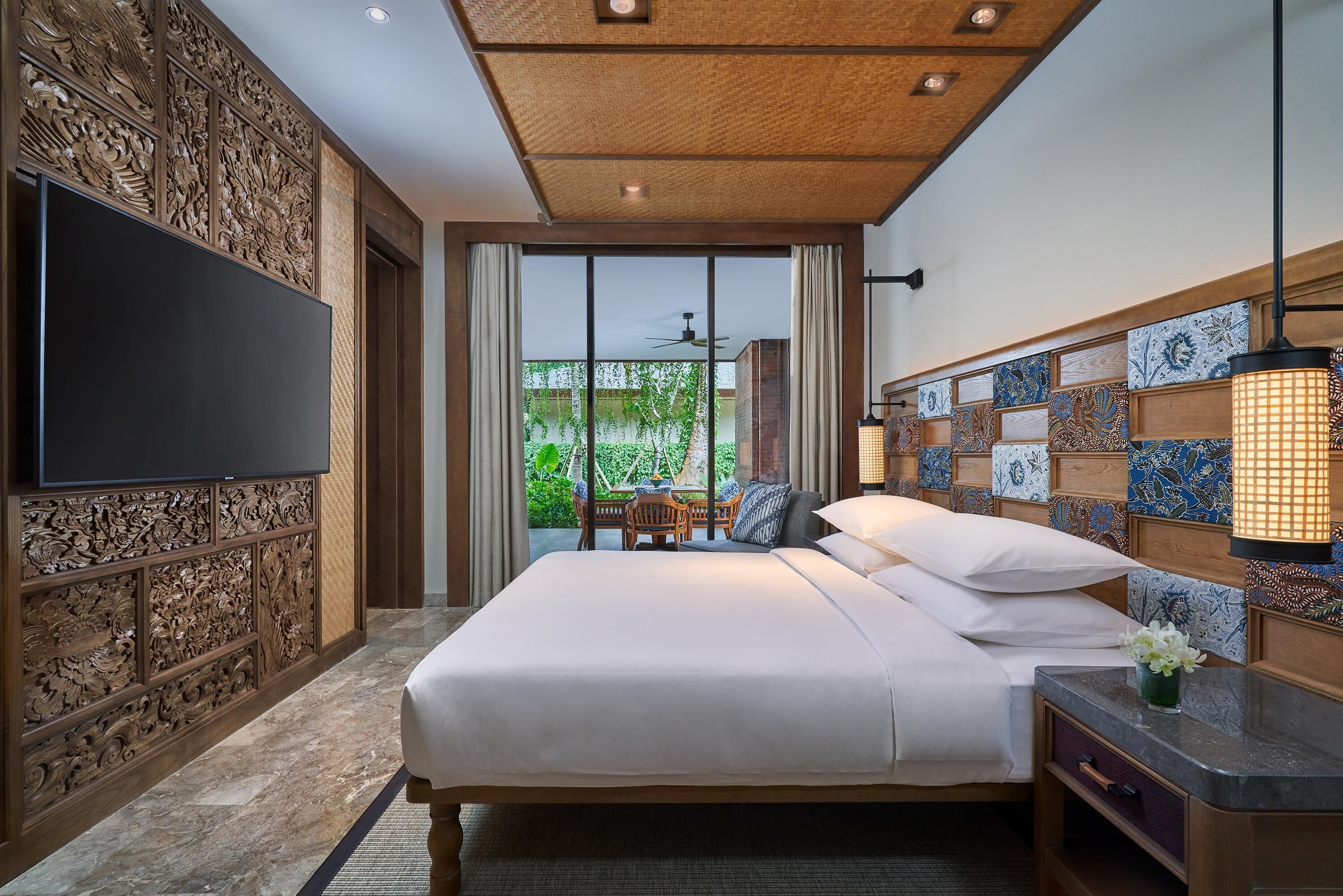 Um quarto amplo com uma cama grande, uma TV de tela plana, uma cabeceira de madeira e vista para um jardim através de uma porta deslizante.