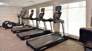 Uma sala de fitness com esteiras e bicicletas ergométricas, grandes janelas e carpete estampado.
