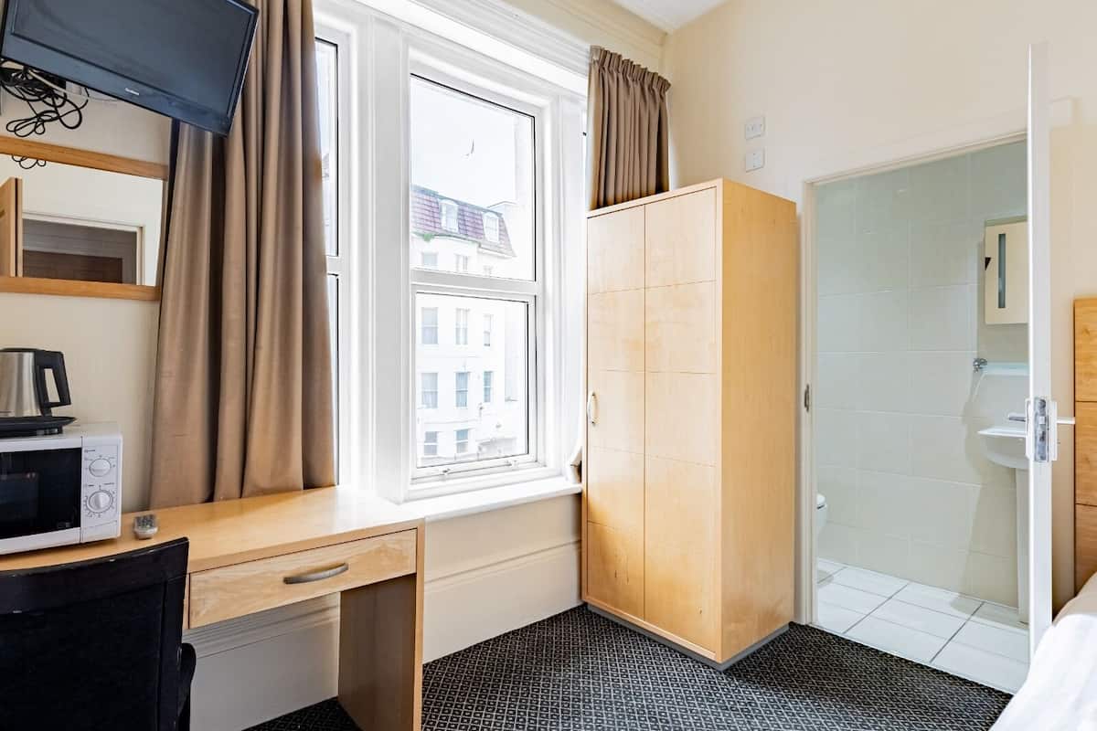 Quarto de hotel com escrivaninha, micro-ondas, armário, área de chuveiro e televisão fixada na parede.