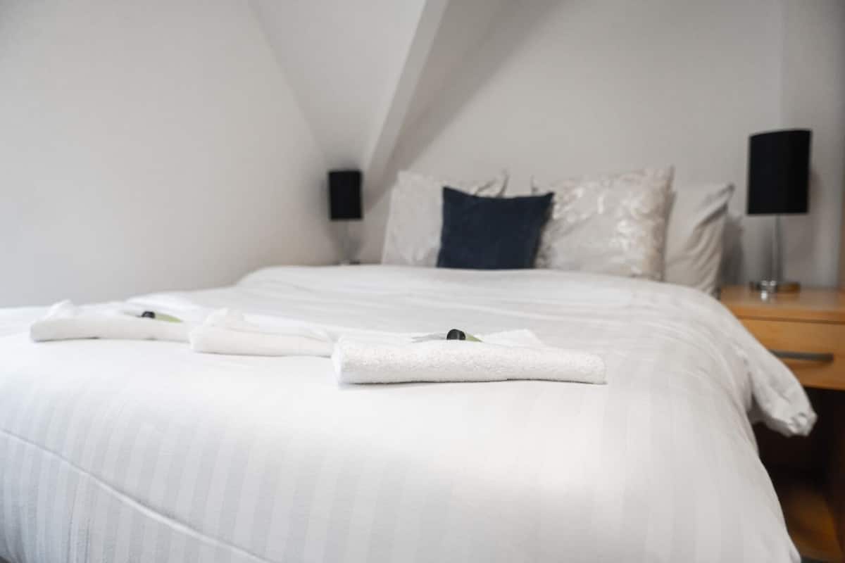 Cama bem arrumada com lençóis e toalhas brancas, mesa de cabeceira de madeira e duas arandelas pretas na parede.