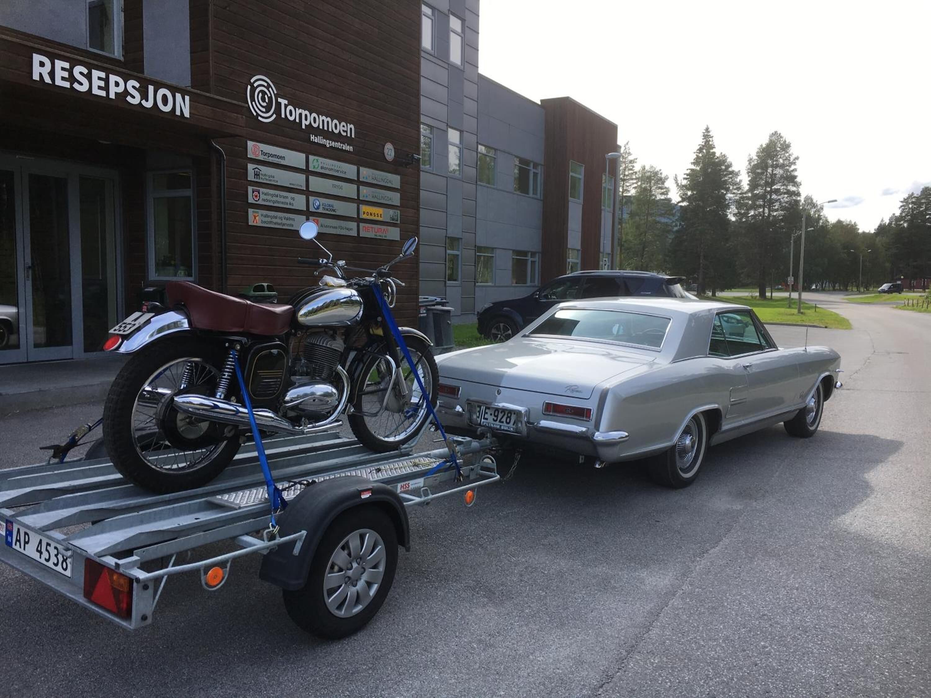 Uma motocicleta e um carro clássico em um reboque, em frente a um edifício com uma placa indicando 'Torpoen Hallingenslenn'.