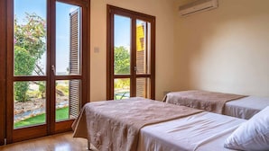 Quarto com duas camas, janelas amplas com persianas, vista para o verde lá fora e ar condicionado na parede.