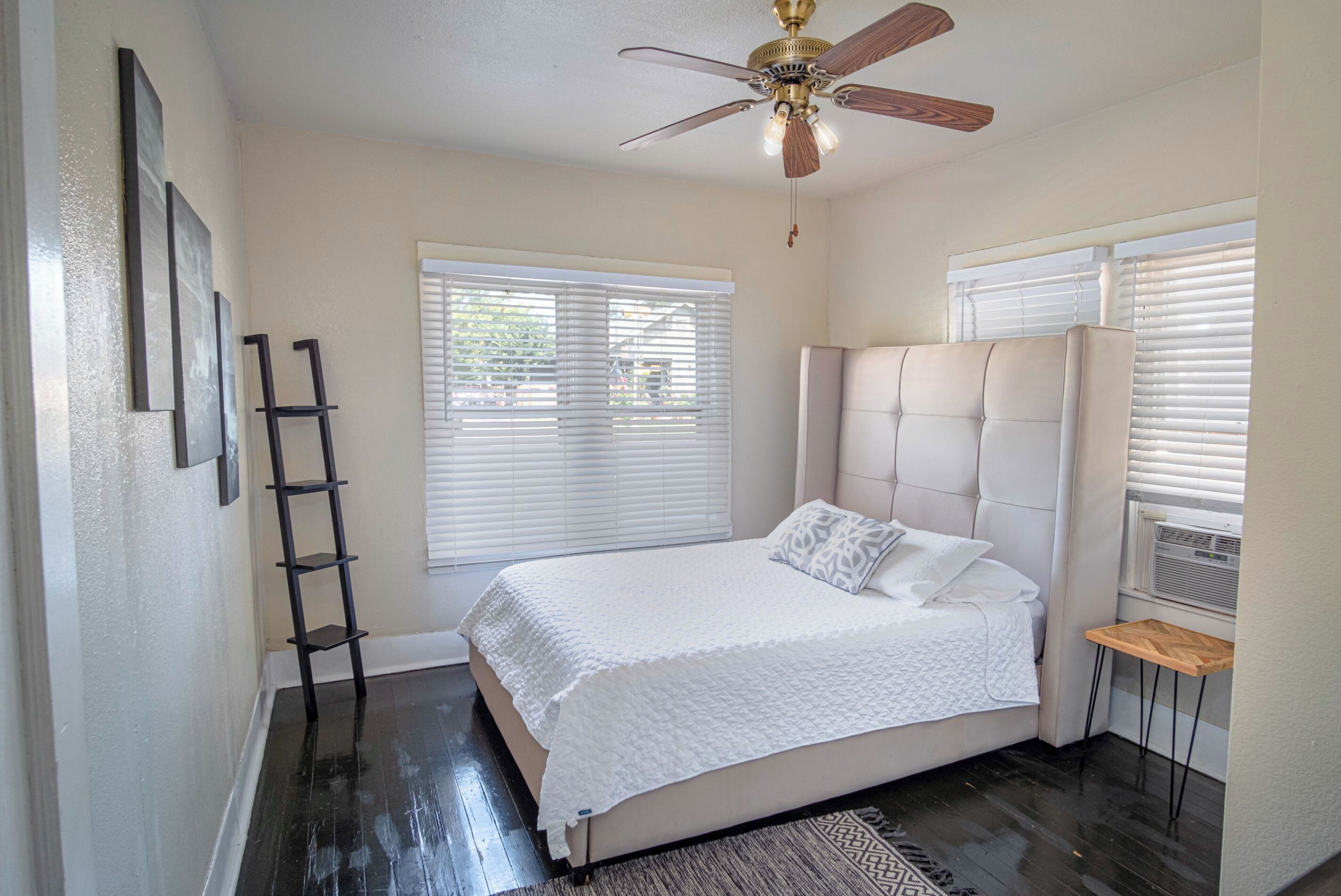 Quarto com cama, ventilador de teto, janela com persianas e uma mesinha.