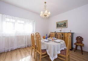 Uma área de refeições com mesa posta para quatro, cadeiras de madeira, um candelabro, um quadro na parede e um radiador sob uma cortina transparente.